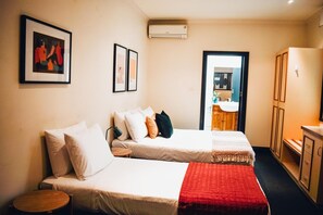 Quarto de hotel com duas camas, um cobertor vermelho, uma pequena mesa redonda e um guarda-roupa.