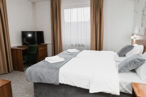 Quarto de hotel com cama, escrivaninha com televisão e janela com cortinas.