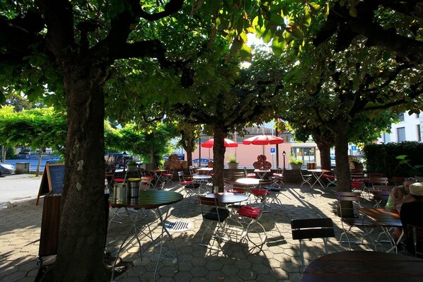 Área de café ao ar livre, com mesas e cadeiras, cercada por árvores.