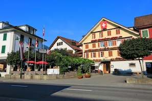 Um hotel com arquitetura europeia tradicional, exibindo uma cruz vermelha proeminente na fachada, diversas janelas com persianas e uma placa indicando ser um hotel.