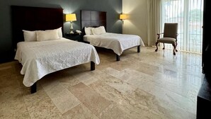 Quarto de hotel com duas camas, uma cadeira, uma escrivaninha e uma janela grande.