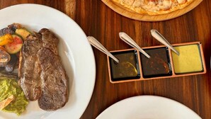 Mesa de madeira com diversos pratos, incluindo massa, pizza, bife e salmão, além de acompanhamentos.