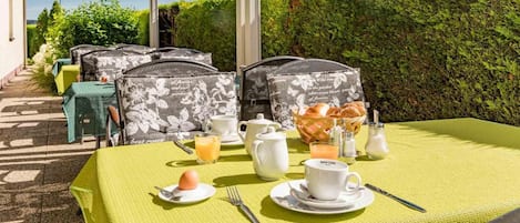 Área de refeições ao ar livre com mesas preparadas para o café da manhã, apresentando toalha de mesa verde, louça branca e uma cesta de pão.