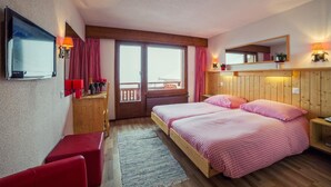 Quarto com cama de casal, cómoda de madeira, um banco vermelho, televisão e varanda com guarda-corpo de madeira.