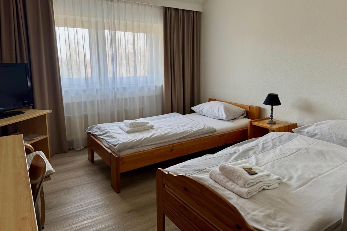 Quarto de hotel com duas camas de madeira, uma escrivaninha com televisão, uma luminária e uma janela com cortinas.