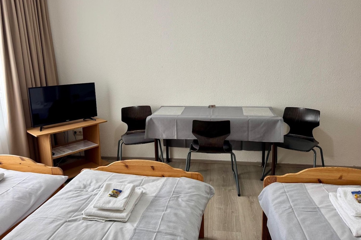 Quarto de hotel com televisão, uma mesa com toalha, quatro cadeiras e duas camas com roupa branca.