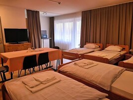 Quarto de hotel com quatro camas de solteiro, televisão, mesa com cadeiras e cozinha compacta ao fundo.