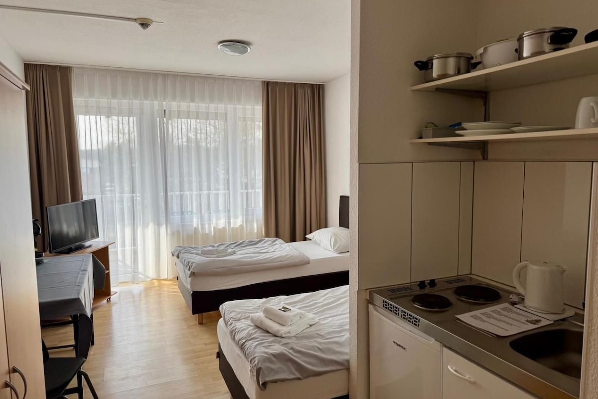 Quarto de hotel com cama, escrivaninha com televisão, cozinha compacta com fogão e pia, e janela com cortinas.
