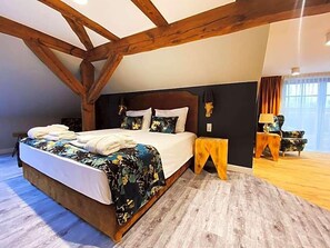 Quarto com uma cama grande, vigas de madeira, mesa de cabeceira, abajur e vista para o exterior.
