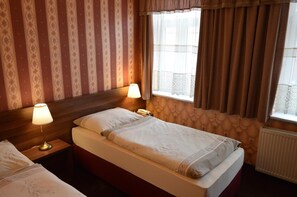 Quarto de hotel com uma cama de solteiro, mesa de cabeceira com abajur e janela com cortinas.