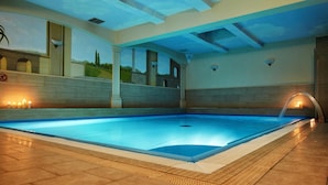 Piscina interior com piso em azulejo, uma fonte de água e vista para o exterior através de grandes janelas.