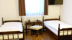 Quarto de hotel com duas camas de solteiro, piso de madeira, escrivaninha com cadeira, janela com cortinas e TV de tela plana na parede.