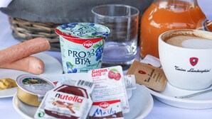 Mesa de café da manhã com pães, jarra de suco de laranja, xícara de café e diversos patês e acompanhamentos.