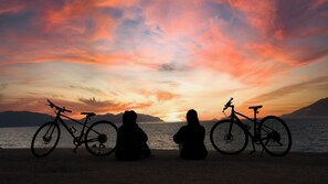 Pôr do sol com silhuetas de duas pessoas e duas bicicletas na praia.