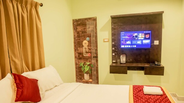 Quarto de hotel com cama, televisão na parede e uma pequena estante com uma planta.