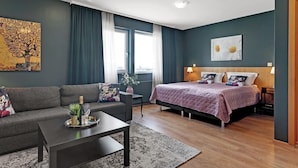 Quarto de hotel moderno com sofá cinza, mesa de centro de madeira, cama com colcha rosa e uma janela grande com cortinas.