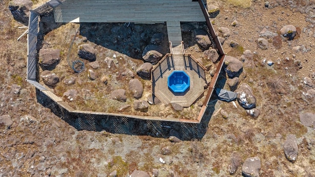 Estrutura hexagonal de madeira com uma banheira de hidromassagem azul central, cercada por uma área com pedras e vedação.