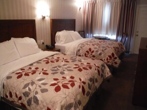 Quarto de hotel com duas camas, cada uma com colcha floral, cabeceira de madeira, mesa de cabeceira com abajur e janela com cortinas.