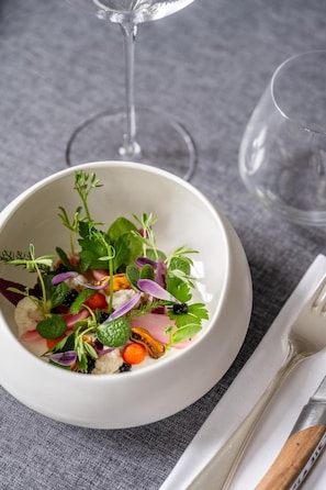 Uma saladeira com diversos vegetais e folhas verdes, acompanhada por uma taça de vinho e um garfo sobre uma mesa.