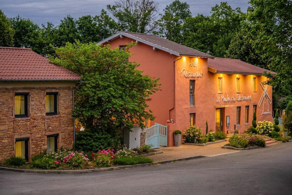 Hotel com fachada vermelha, um edifício de pedra à esquerda e uma entrada bem iluminada.