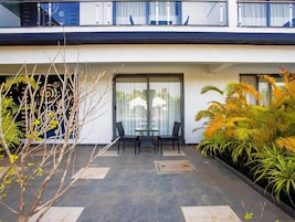 Um edifício moderno com varanda, área de pátio com mesa e cadeiras, e plantas tropicais.