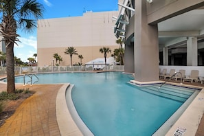 Piscina com borda curva, rodeada por palmeiras e um edifício ao fundo.