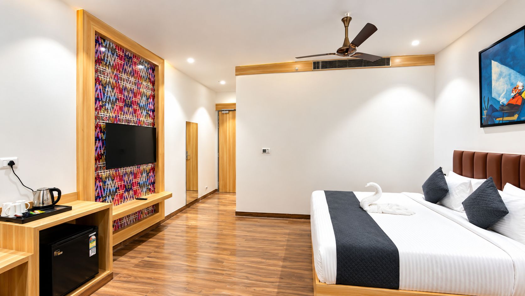 Quarto de hotel com cama, televisão, secretária com chaleira, ventoinha no teto e decoração colorida na parede.