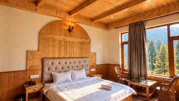 Quarto com painéis de madeira, uma cama grande, mesa de cabeceira, abajur e vista para árvores pela janela.