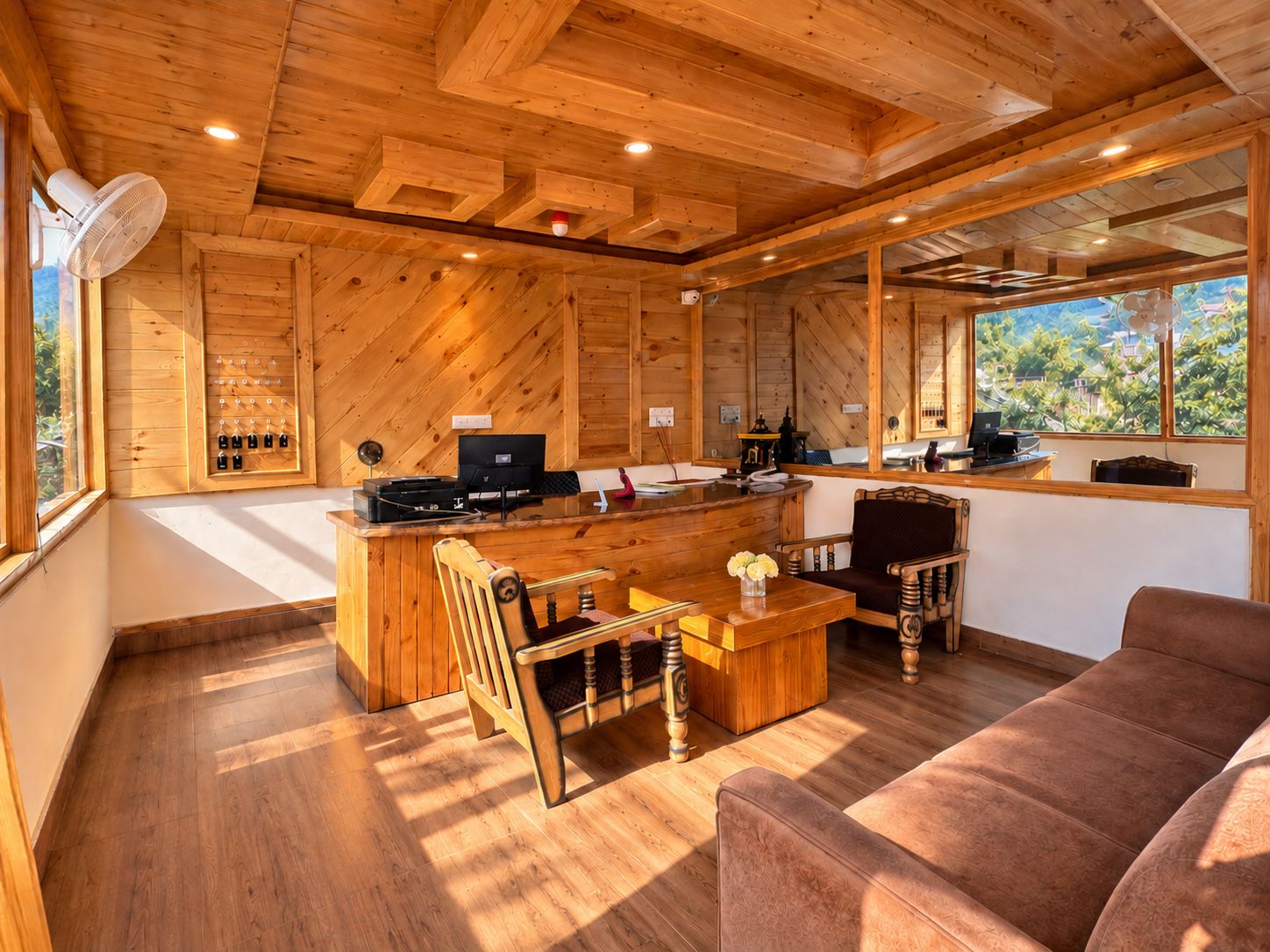 Interior de uma cabana de madeira com escrivaninha, cadeiras, sofá e uma grande janela com vista para o exterior.