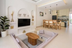 Uma sala de estar moderna com televisão de tela plana, uma mesa de centro de madeira e um sofá. Há uma área de jantar com cadeiras de madeira e uma cozinha ao fundo.