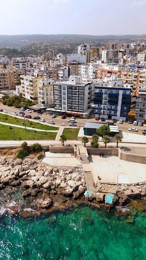 Uma área urbana costeira com um edifício notável identificado como 'KORASION' e um litoral rochoso com águas cristalinas.