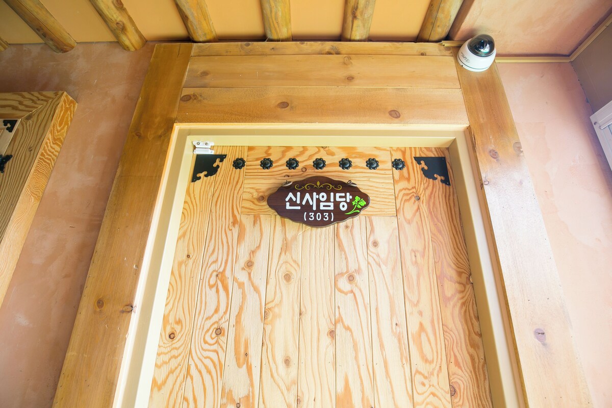 Uma porta de madeira com uma placa que diz '안녕하세요 (303)', indicando uma saudação e um número de quarto.