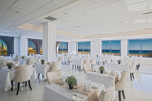 Uma ampla área de jantar com mesas preparadas para o serviço, grandes janelas com vista para o mar e um design moderno no teto.