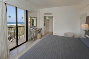 Quarto de hotel com cama, escrivaninha, poltrona, uma grande janela com vista para o oceano e uma varanda.