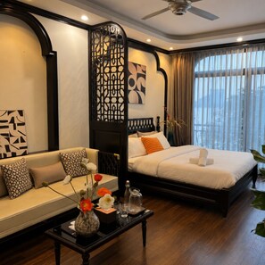 Quarto de hotel com sofá, cama, biombos decorativos, ventoinha de teto e janela com cortinas.
