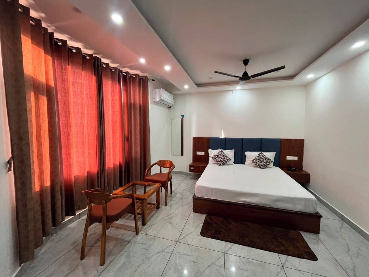 Quarto de hotel com cama, escrivaninha, cadeiras, ventoinha no teto e ar condicionado.