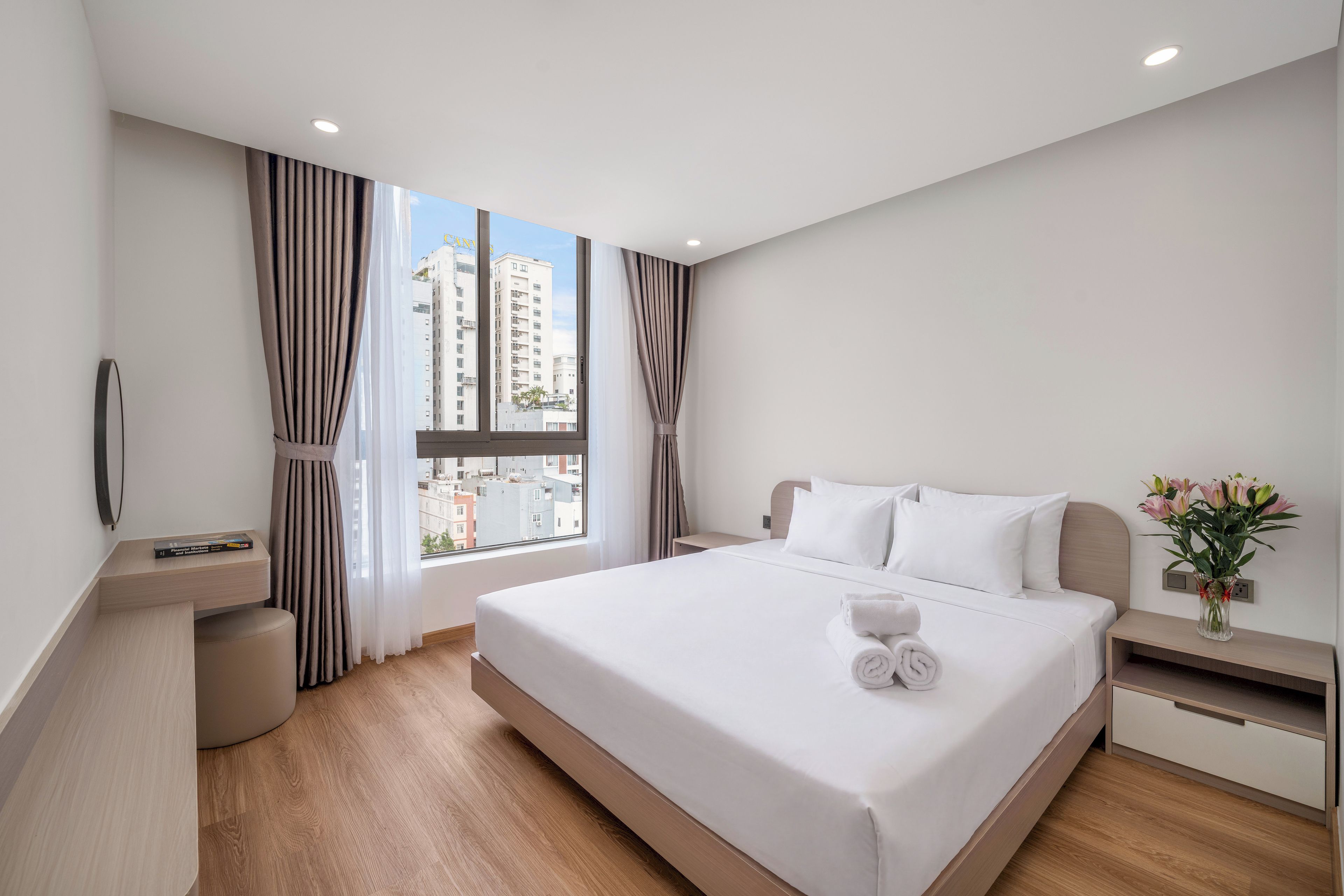 Quarto de hotel com uma cama grande, mesa de cabeceira, cortinas e vista para prédios.