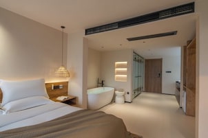 Quarto de hotel moderno com uma cama grande, mesa de cabeceira, luminária pendente, banheira e chuveiro separado.