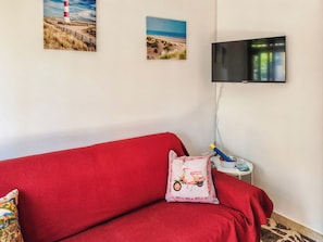Um sofá vermelho com almofada em formato de scooter, uma televisão fixada na parede e dois quadros emoldurados.