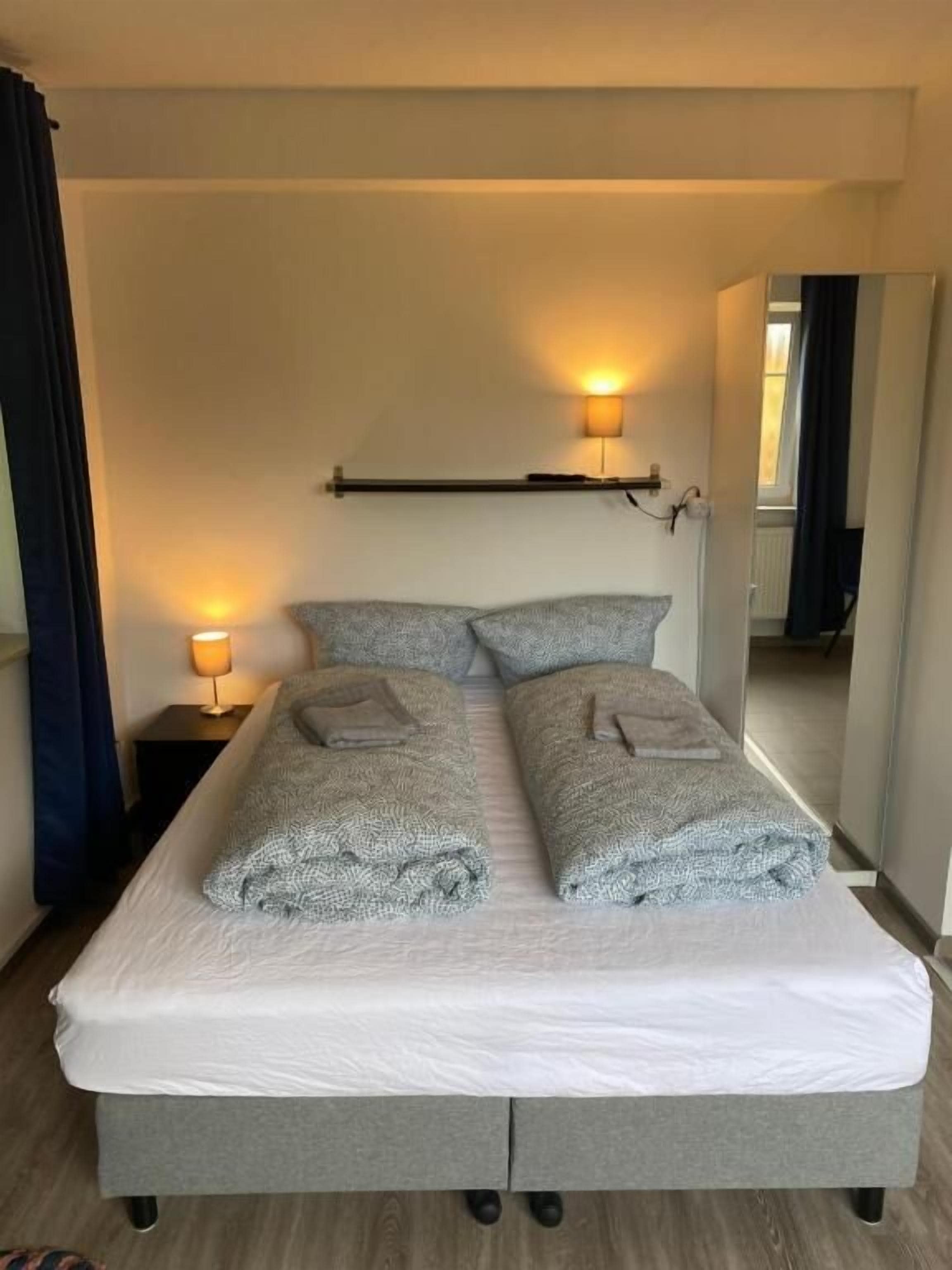 Uma cama de casal com colchão branco e roupa de cama cinza, duas luminárias de cabeceira e um espelho na parede.