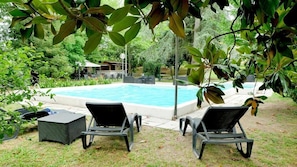 Piscina com espreguiçadeiras e uma pequena mesa, cercada por vegetação.