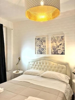 Quarto com cabeceira estofada, cama com roupa de cama branca, mesa redonda com abajur e duas obras de arte penduradas na parede.