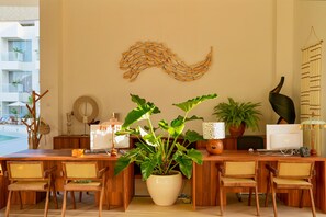 Quarto com escrivaninha de madeira, duas cadeiras, uma grande planta em vaso, um objeto decorativo na parede e vista para um edifício lá fora.