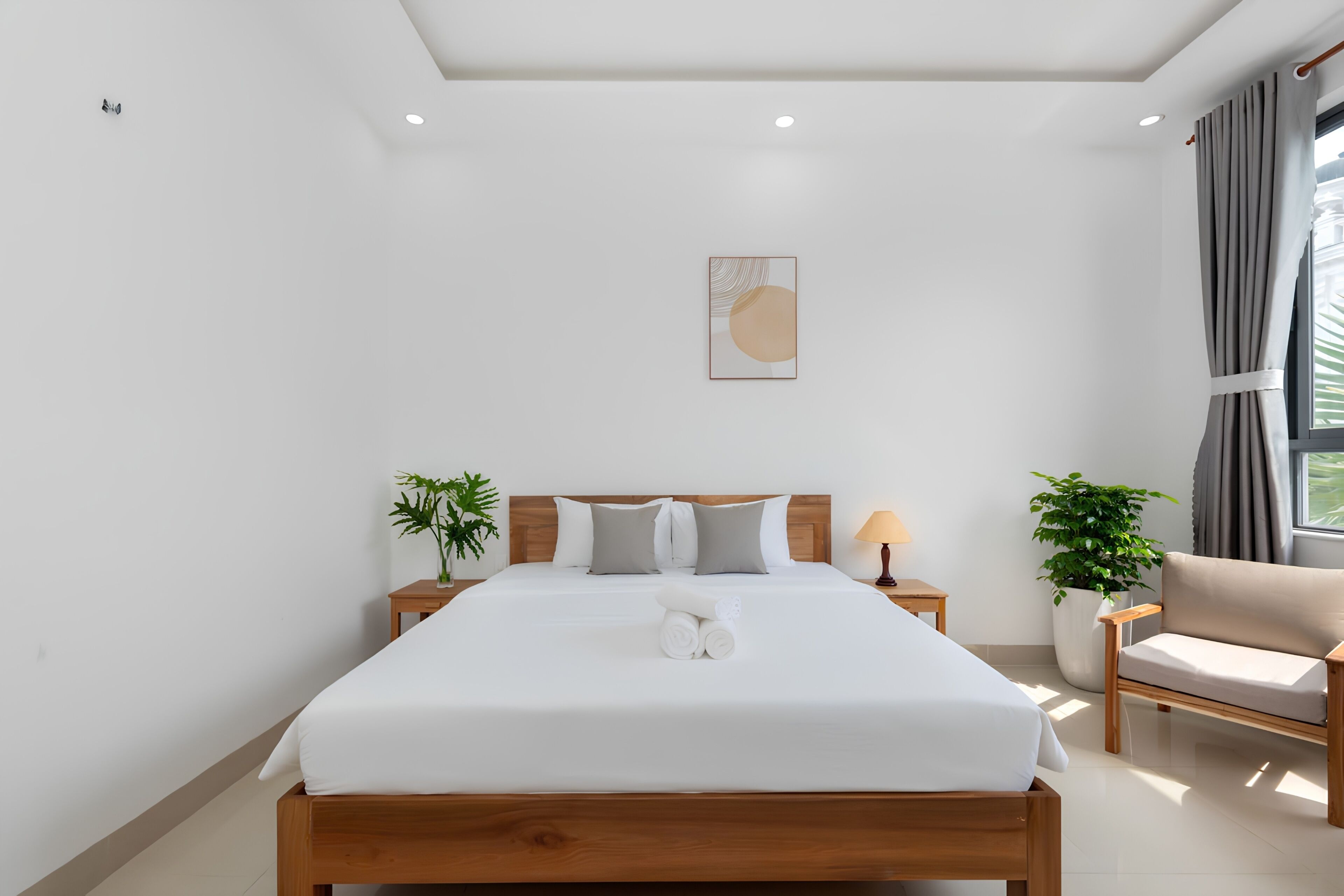 Quarto moderno com cama de madeira, roupa de cama branca, duas mesas de cabeceira com abajures, uma cadeira e um vaso com planta.