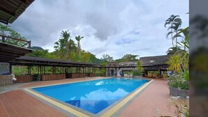 Uma piscina retangular com um deck revestido ao redor, uma área de estar coberta à esquerda e um edifício com telhado de palha à direita.