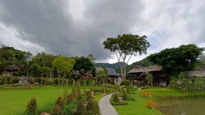 Um caminho serpenteia por um jardim bem cuidado, com um lago, cercado por edifícios de estilo tradicional.
