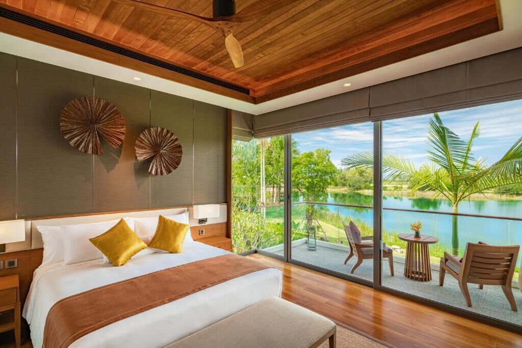 Quarto de hotel moderno com uma cama grande, uma varanda com vista para um lago e um espaço de refeições com cadeiras e mesa.