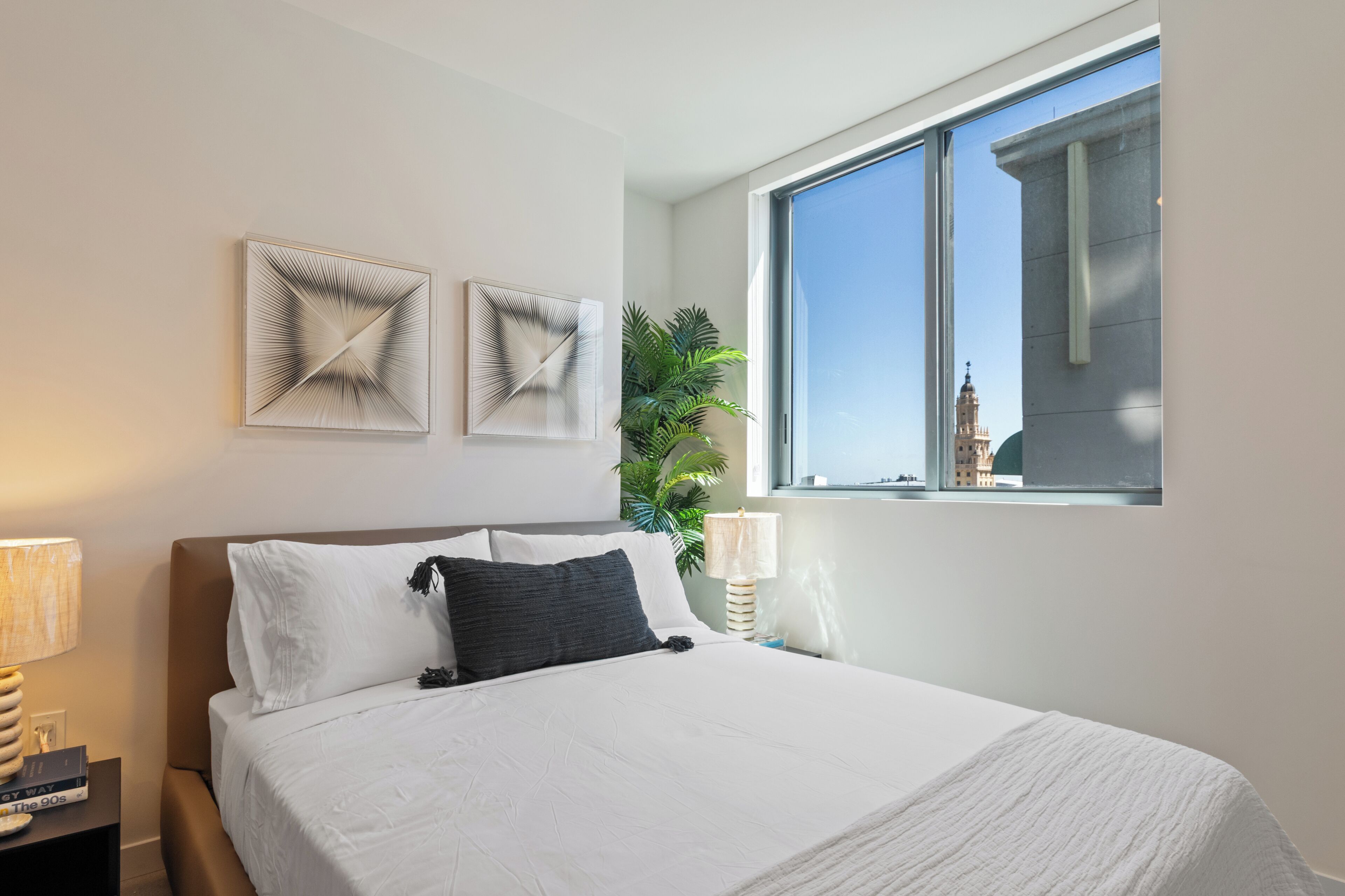 Quarto de hotel com uma cama, duas obras de arte penduradas na parede, uma janela com vista para um edifício e um vaso de plantas.