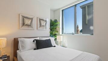 Quarto de hotel com uma cama, duas obras de arte penduradas na parede, uma janela com vista para um edifício e um vaso de plantas.