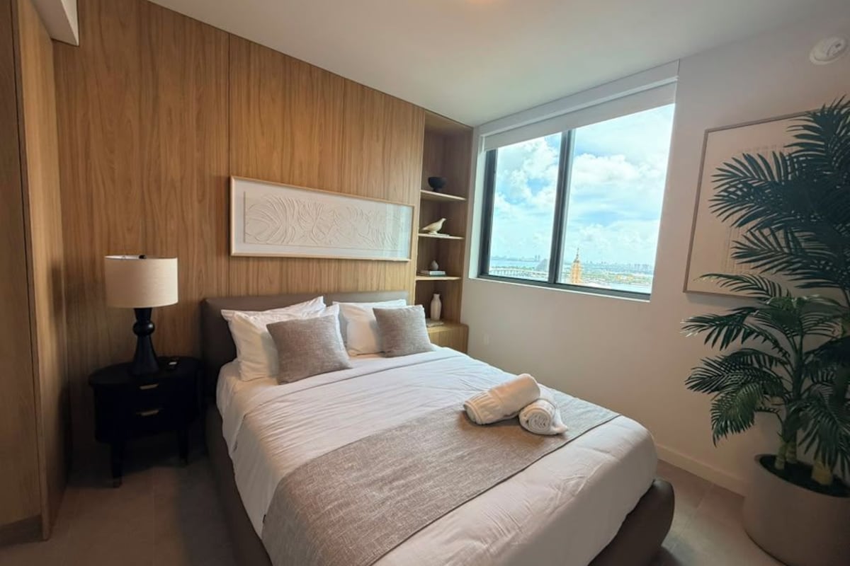 Quarto de hotel com cama grande, mesa de cabeceira, luminária, estante de livros, janela ampla com vista para a cidade e uma planta em vaso.